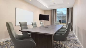 Uma sala de conferências com uma mesa comprida, seis cadeiras, um quadro branco, uma televisão de tela plana e janelas grandes com cortinas.