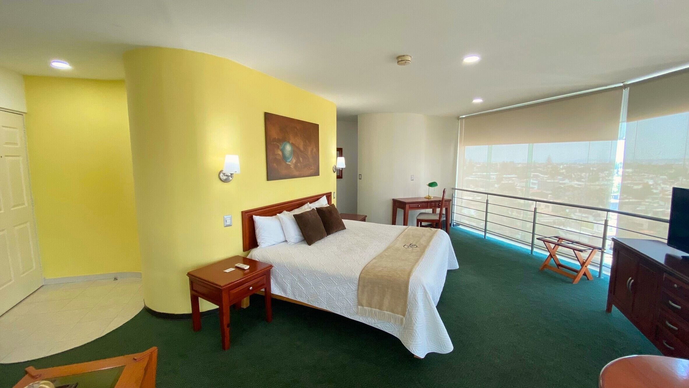 Quarto de hotel com cama, mesas de cabeceira, escrivaninha, cadeira, televisão e varanda com vista para a cidade.