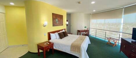 Quarto de hotel com cama, mesas de cabeceira, escrivaninha, cadeira, televisão e varanda com vista para a cidade.