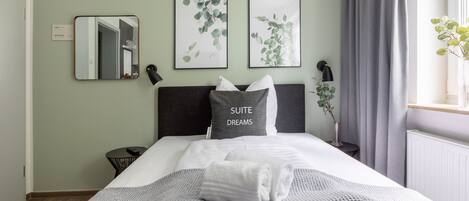 Cama bem arrumada com manta cinza texturizada, travesseiro com a inscrição "SUITE DREAMS" e toalha branca dobrada. Dois quadros botânicos emoldurados na parede, luminária preta e espelho sobre a cama.