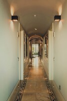 Um corredor comprido e estreito com chão estampado, paredes claras e luminárias fixadas na parede.