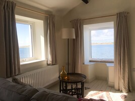 Quarto com cama, uma mesinha com abajur e vista para o mar pela janela.