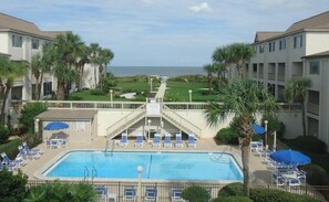 Área de piscina com espreguiçadeiras e guarda-sóis, cercada por edifícios com vista para a praia.