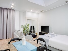 Quarto de hotel moderno com cama, televisão, área de estar com mesa e cadeiras, e um espelho grande.