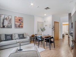 Uma sala de estar moderna com um sofá cinzento, uma mesa de centro e um espaço de jantar com mesa e cadeiras de madeira. Há também uma cozinha com frigorífico e fogão.