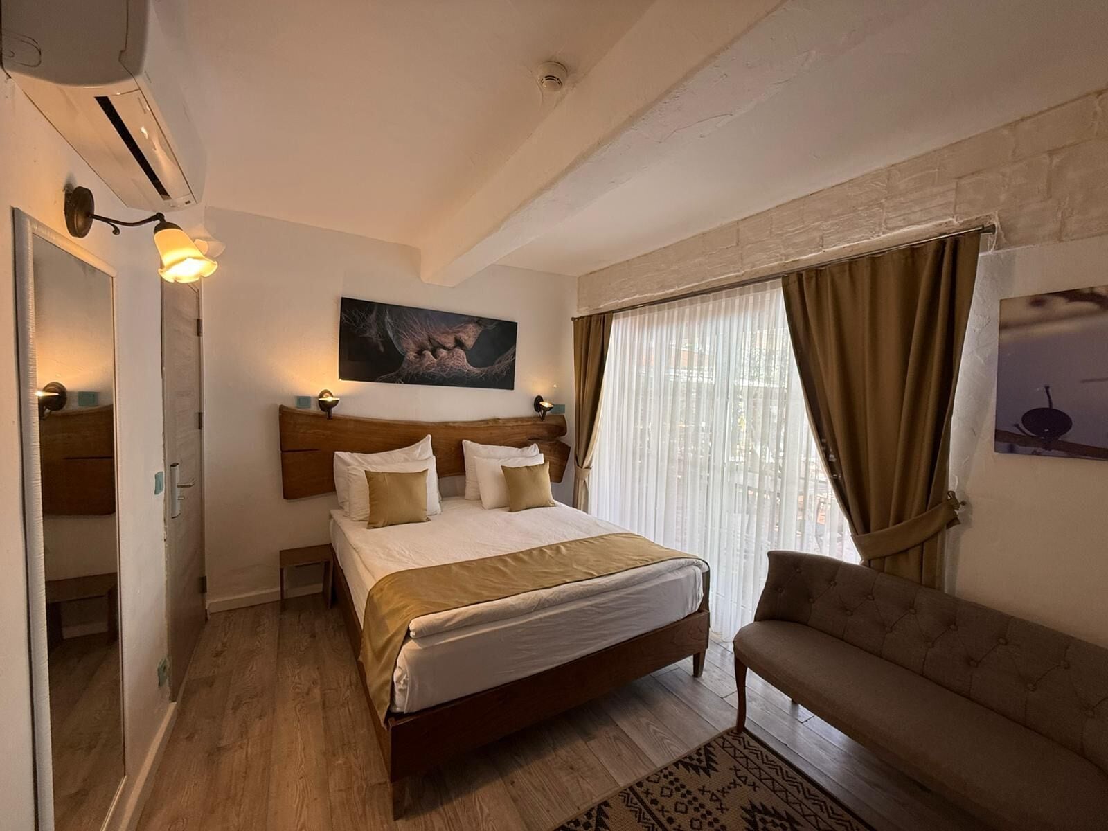 Quarto de hotel com cama de madeira, sofá, pintura na parede e uma grande janela com cortinas.