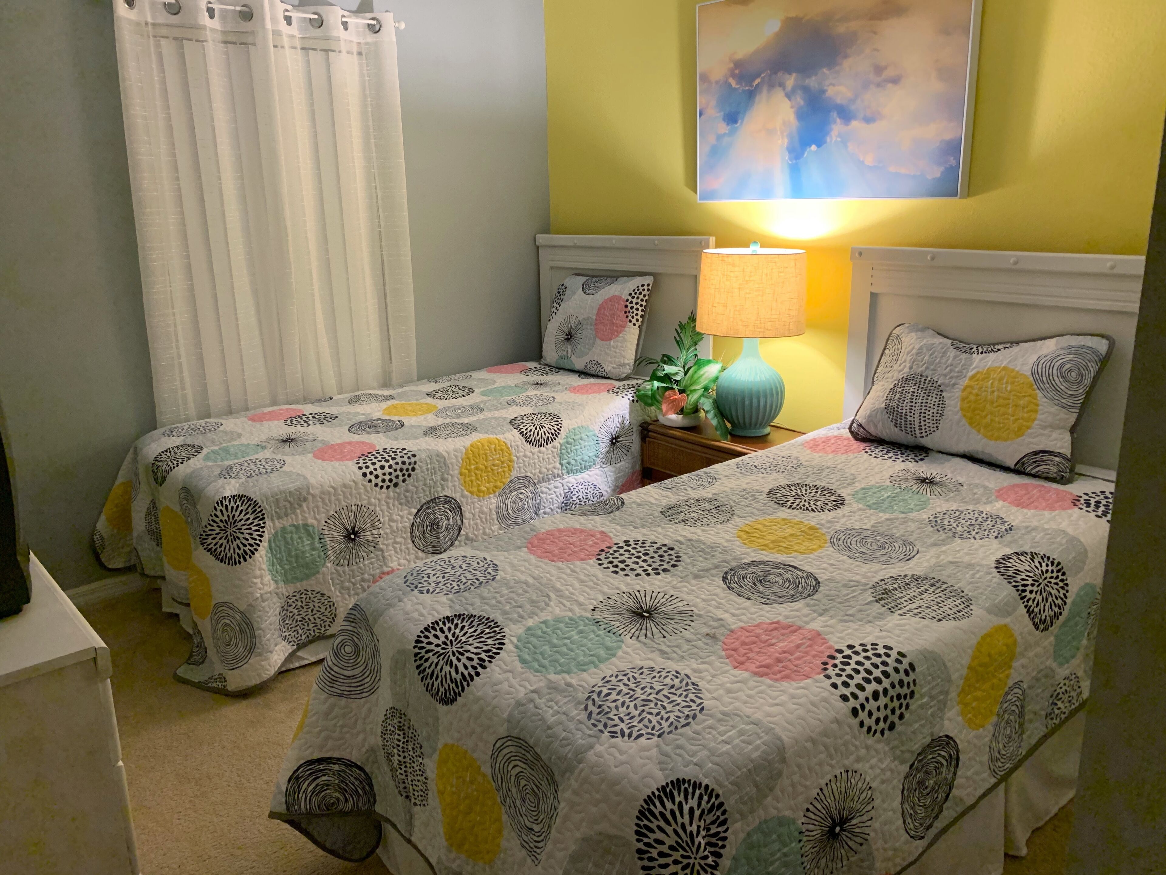 Quarto com duas camas, cada uma com colcha estampada, mesa de cabeceira e luminária, e uma pintura na parede.
