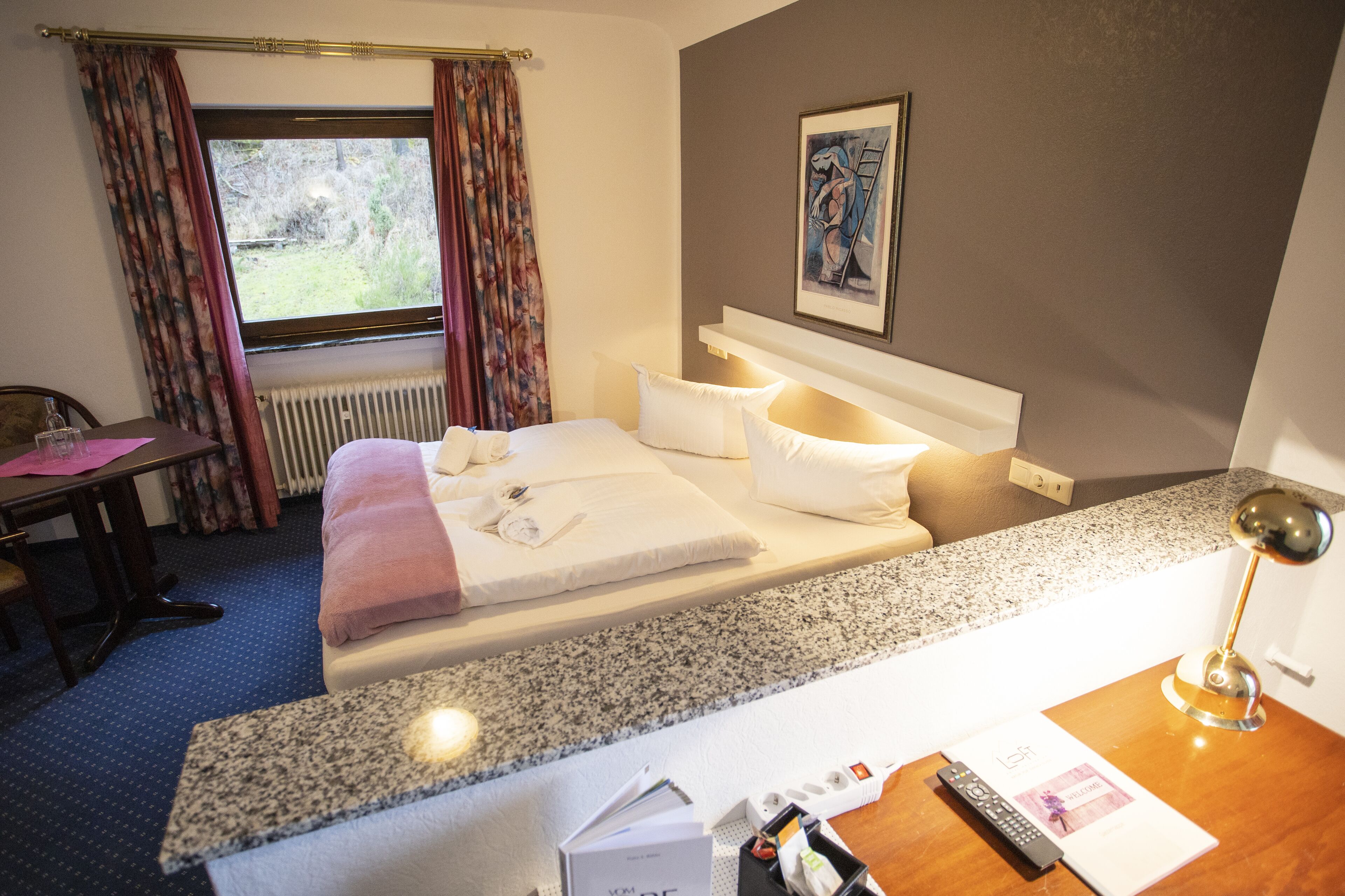 Quarto de hotel com cama, escrivaninha, cadeira, janela com cortinas, radiador, um quadro na parede e abajur na escrivaninha.