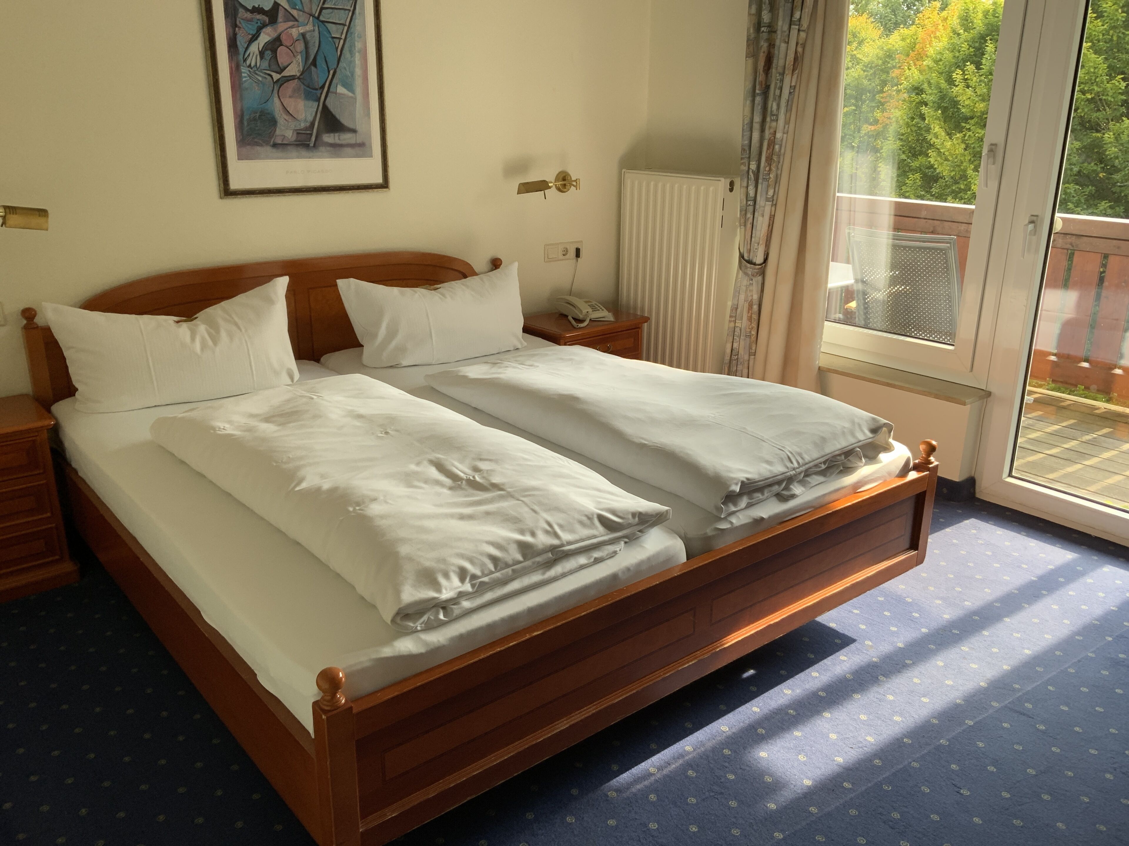 Uma cama de casal com lençóis brancos, cabeceira de madeira, mesa de cabeceira com telefone, radiador e janela com cortinas.