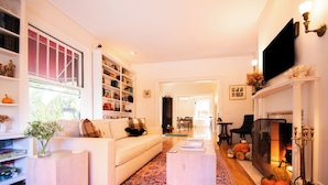 Uma sala de estar com sofá branco, mesa de centro e lareira. Há estantes de livros e uma TV fixada na parede.