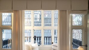 Quarto com um grande janelão com vários painéis, um radiador embaixo e um assento à janela com almofadas e uma revista.
