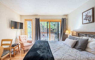Quarto de hotel com cama, televisão, cadeira, escrivaninha e vista para o exterior por uma porta de correr.