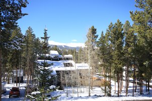 Uma cabana com telhado coberto de neve, rodeada por altas árvores de pinheiro, e uma montanha ao fundo.