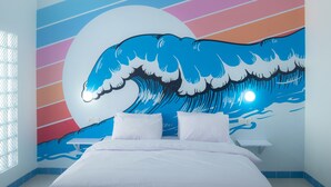 Quarto com um grande mural de ondas na parede, uma cama com roupa de cama branca e uma janela com padrão quadriculado.