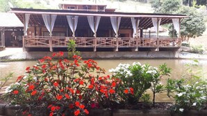 Um edifício à beira do rio com um alpendre coberto e um pequeno jardim com flores vermelhas e brancas em frente.