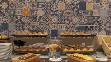Buffet com diversos pães e sobremesas, incluindo croissants e bolos, apresentado em bandejas e num recipiente de vidro.