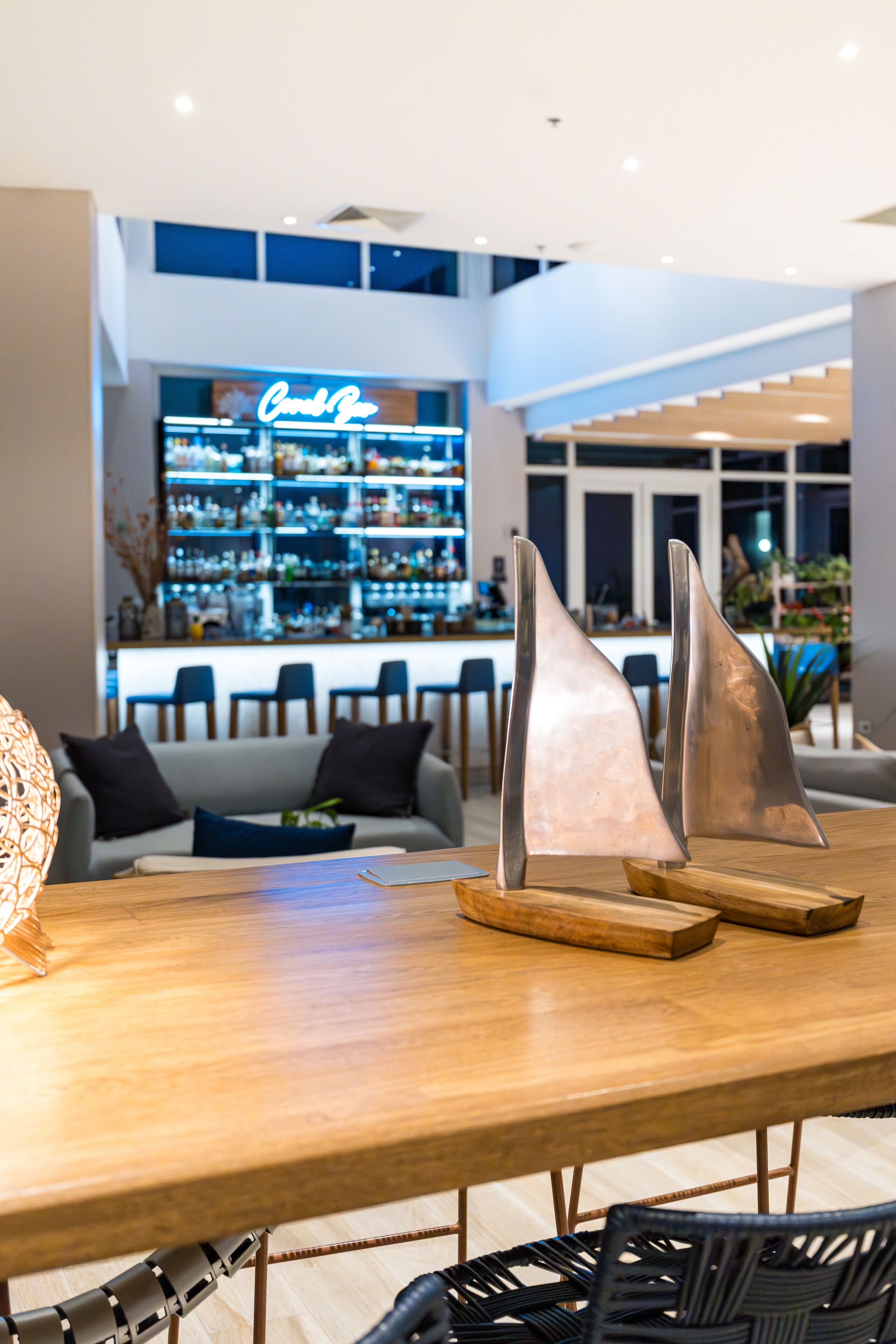 Uma área de bar moderna com um letreiro de neon, uma mesa de madeira com decoração de barcos à vela e um espaço de estar com sofá e cadeiras.