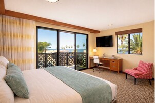 Quarto de hotel com uma cama grande, uma escrivaninha, uma cadeira, uma televisão e varanda com vista para palmeiras e o oceano.