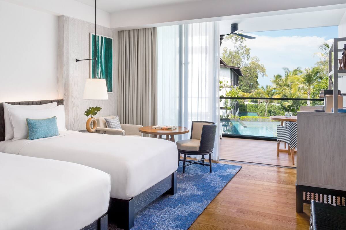 Quarto de hotel com duas camas, uma mesinha, uma cadeira e varanda com vista para a piscina.