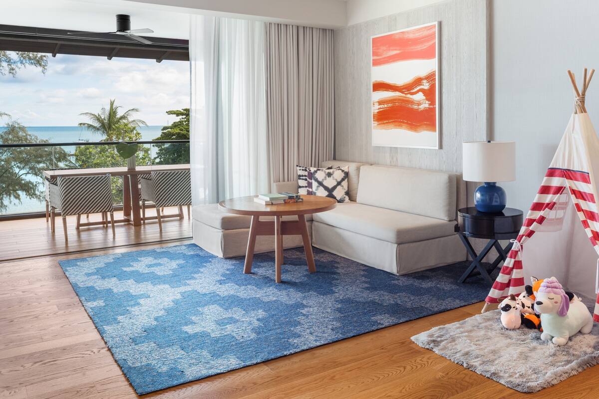 Uma sala de estar moderna com um sofá, uma mesa de centro redonda, um tapete azul estampado, um tipi branco e vista para o oceano.