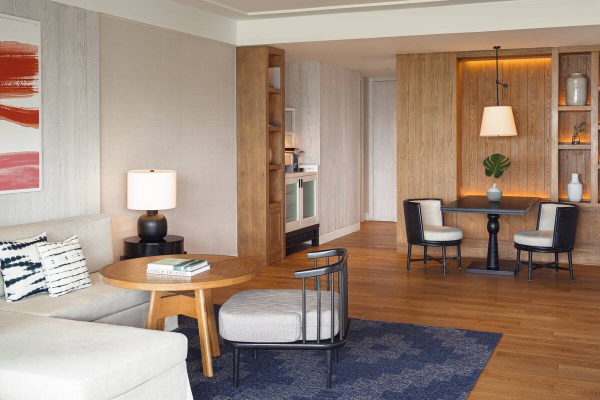 Quarto de hotel moderno com sofá, mesa redonda de madeira, duas cadeiras, estante de livros e luminária.