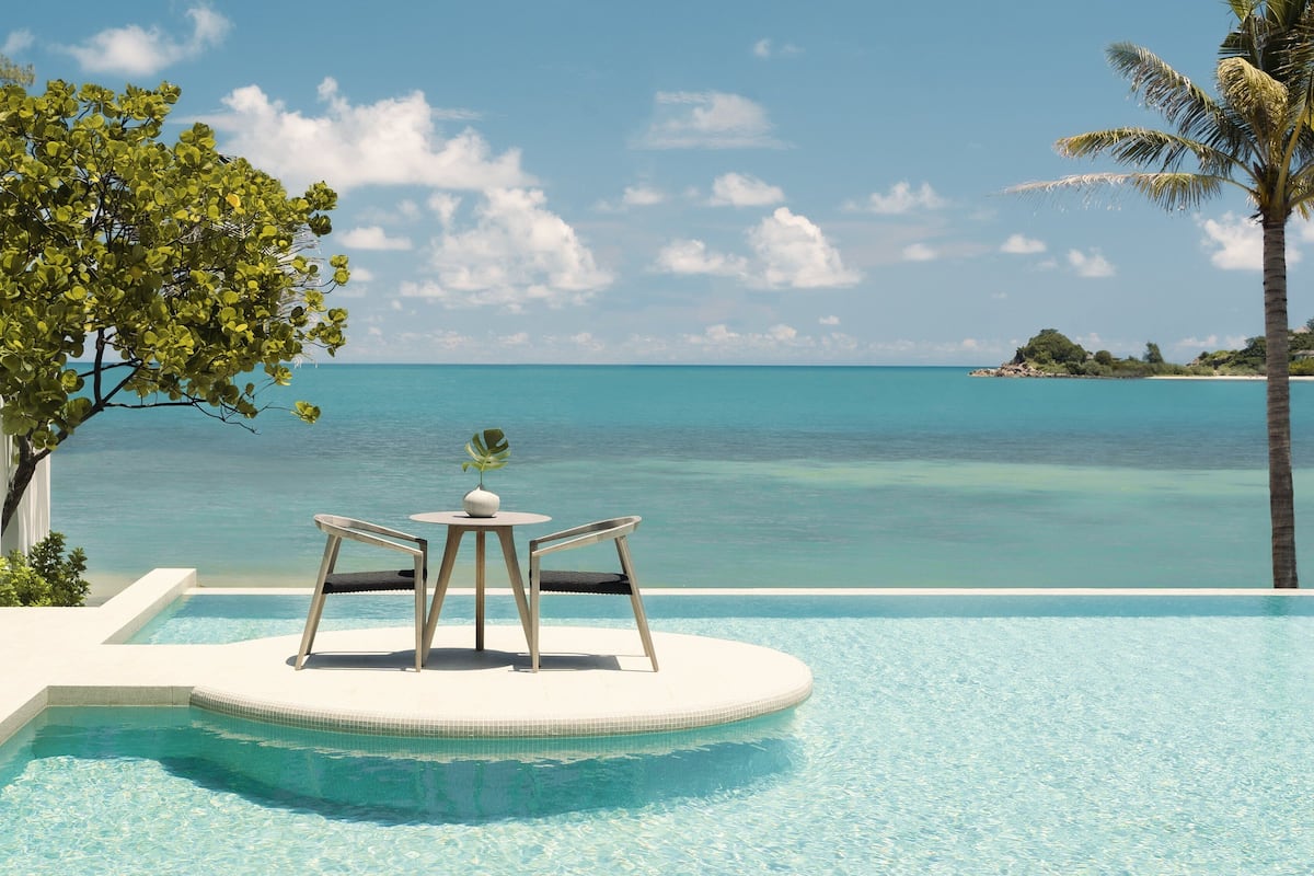 Piscina com duas cadeiras e uma mesa, com vista para um mar azul e uma ilha tropical.