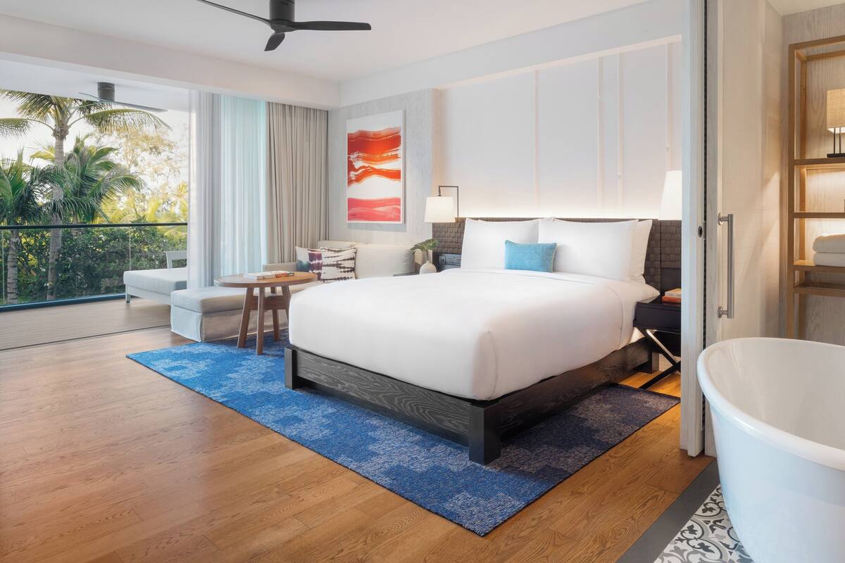Quarto de hotel moderno com uma cama grande, área de estar com uma mesinha, banheira e vista para palmeiras através de janelas amplas.