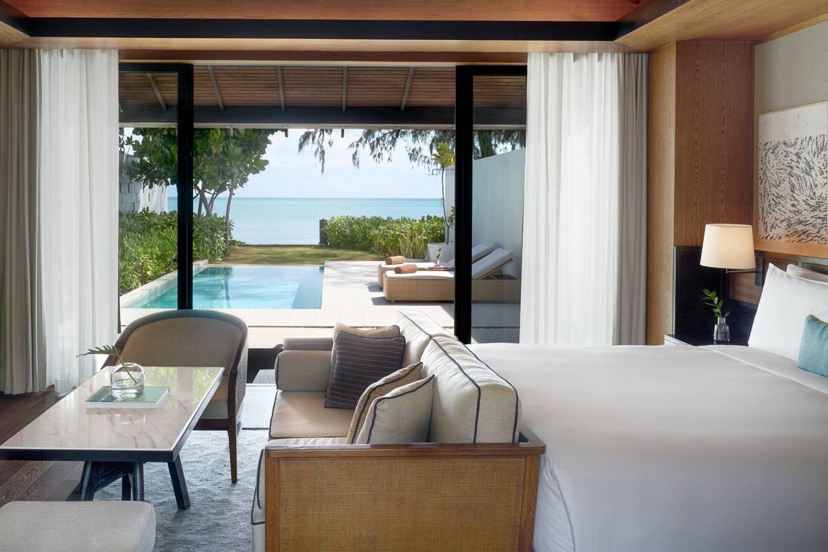 Quarto de hotel com uma cama grande, área de estar com vista para o mar, mesa de jantar e estante de livros.