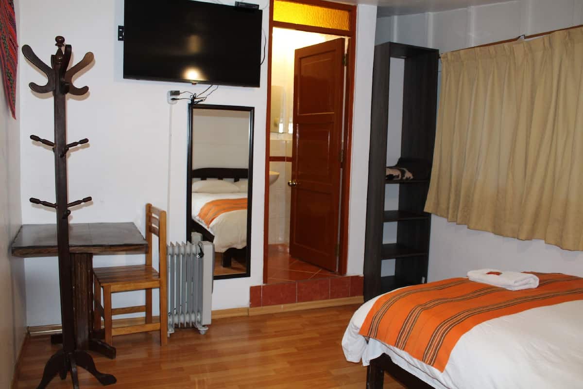 Quarto com cama, uma cadeira de madeira, cabideiro, radiador, televisão e porta que dá para outro cômodo.