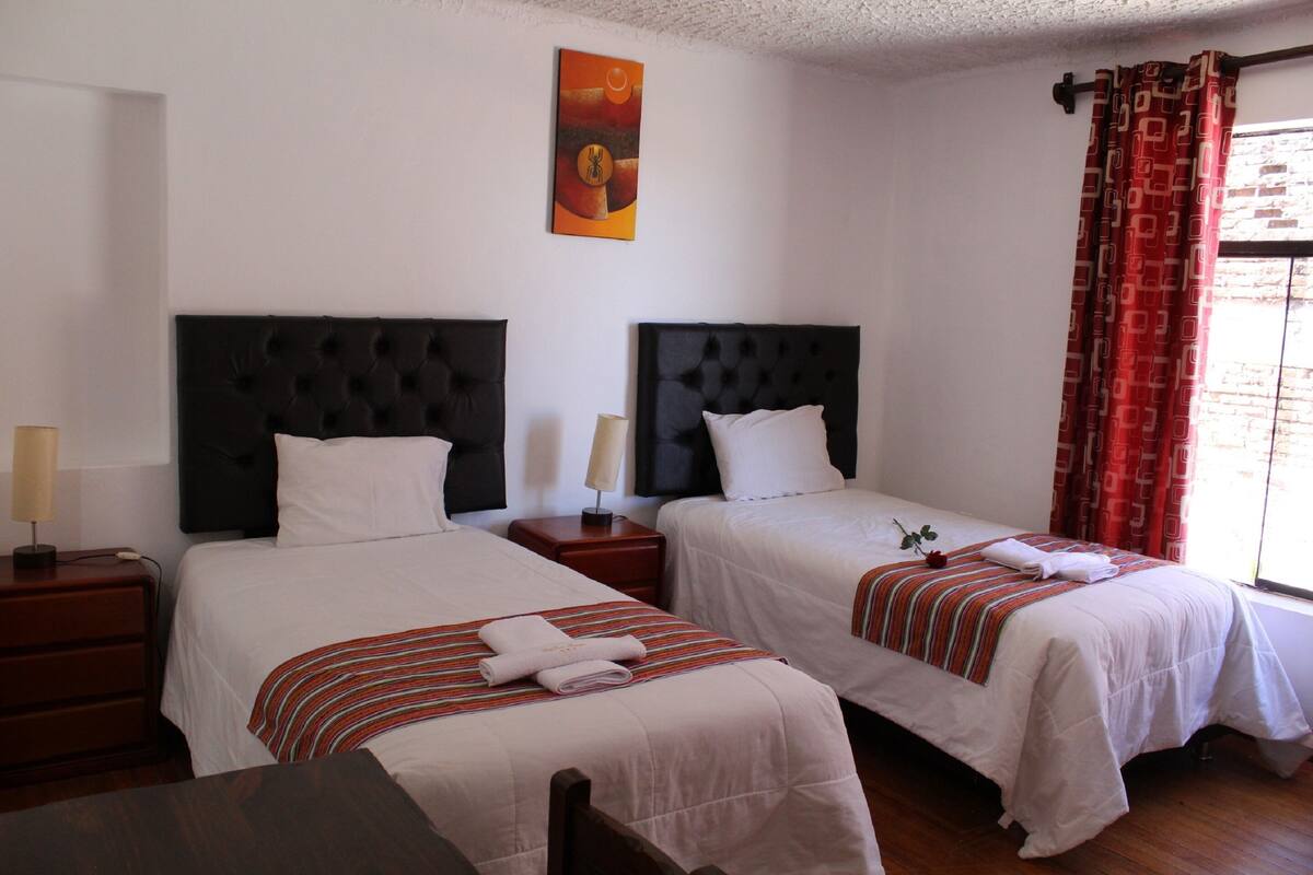 Quarto de hotel com duas camas, cada uma com cabeceira, mesas de cabeceira e uma janela com cortinas.