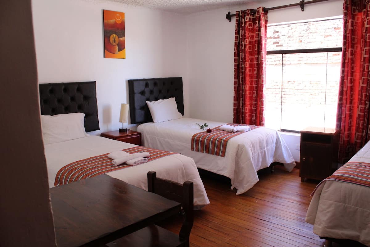 Quarto de hotel com duas camas, piso de madeira, uma mesa e uma janela com cortinas.