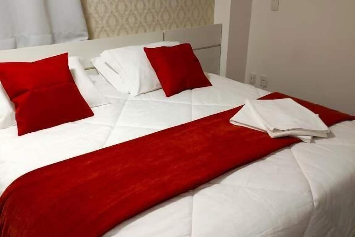 Cama bem arrumada com roupa de cama branca e detalhes em vermelho, incluindo almofadas e um cobertor.