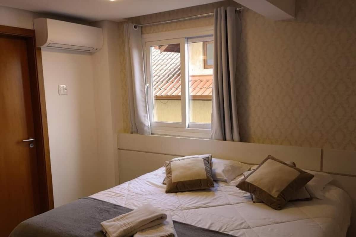 Cama bem arrumada com colcha cinza e dois travesseiros, uma janela com cortinas e uma porta à esquerda.