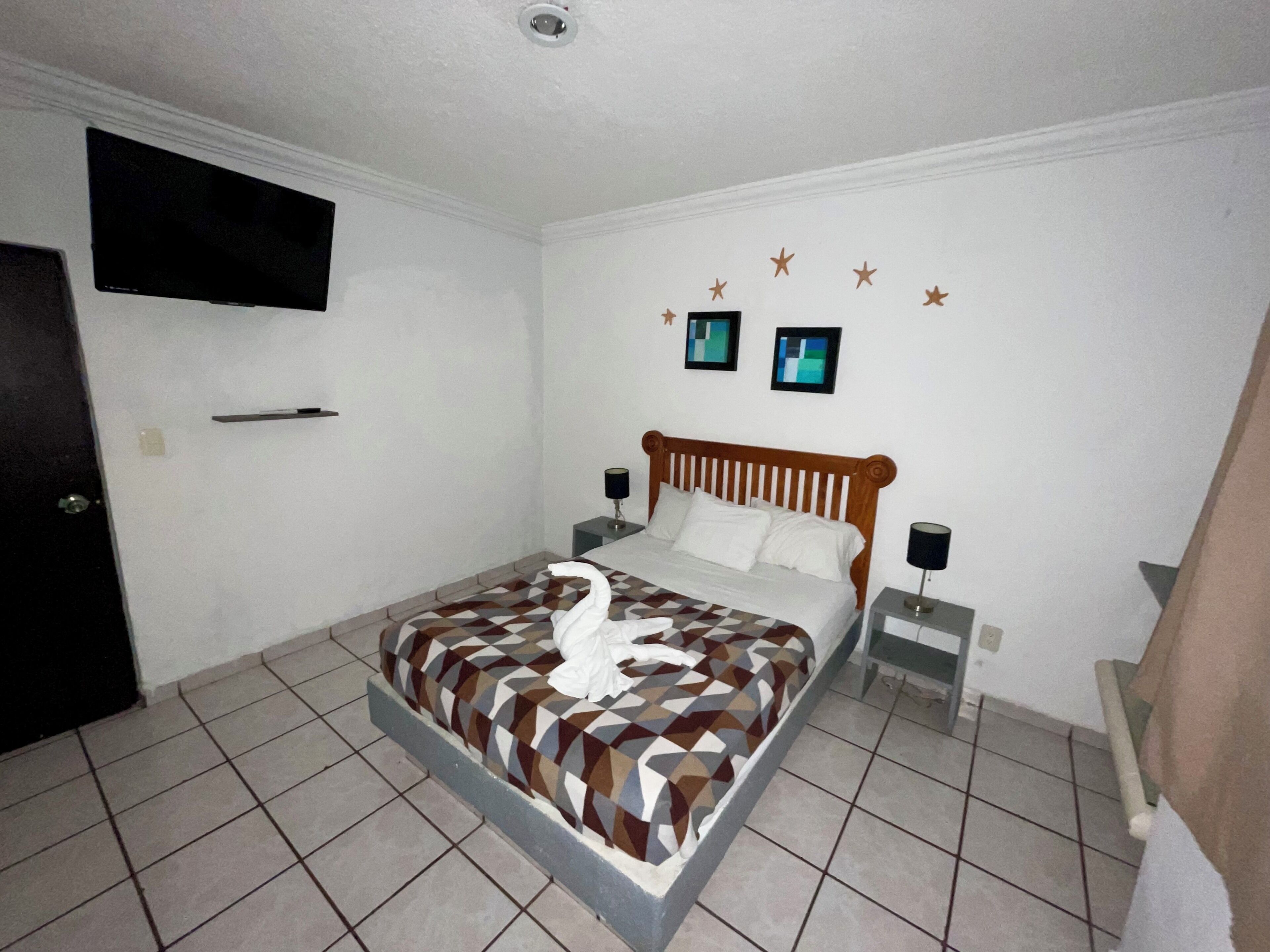 Quarto com cama, duas mesas de cabeceira, televisão na parede e uma porta.