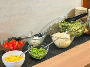 Mesa de buffet com diversos alimentos, incluindo tomates, milho, ervilhas e uma salada.