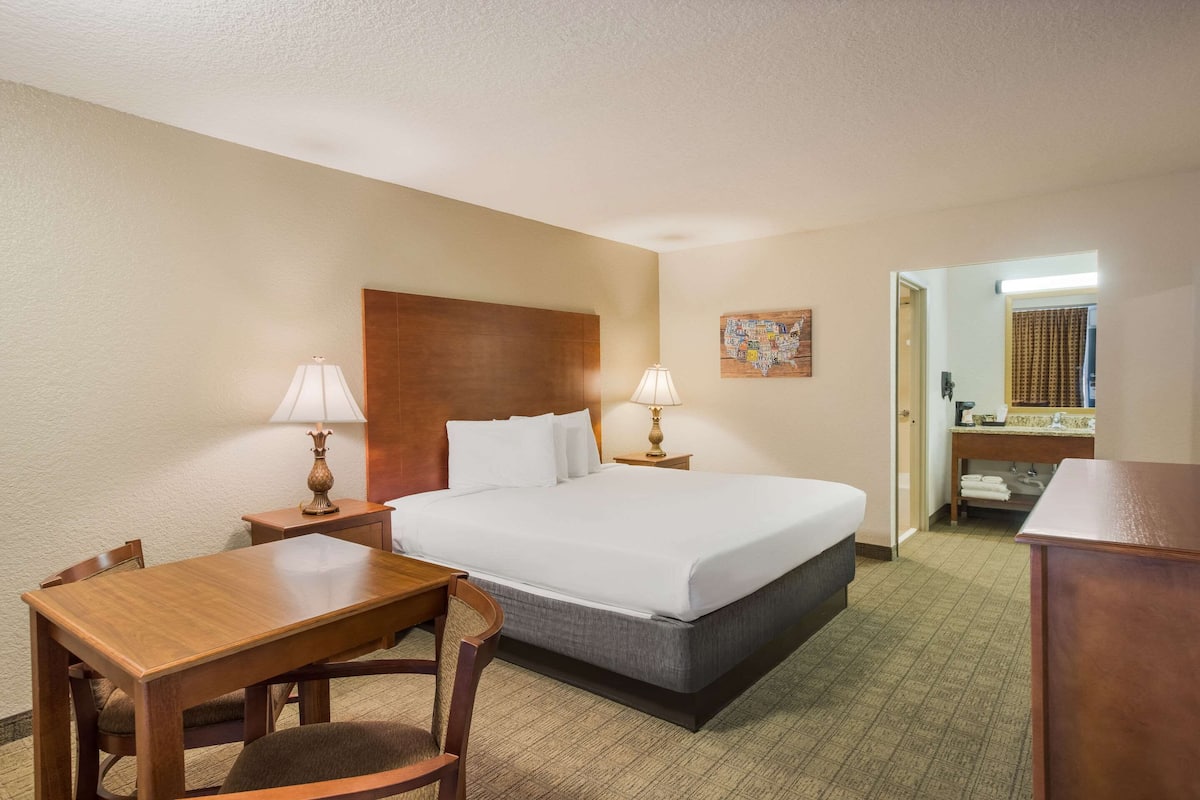 Quarto de hotel com cama, escrivaninha, cadeira, duas luminárias de cabeceira, mesa pequena, espelho e banheiro visível por uma porta aberta.