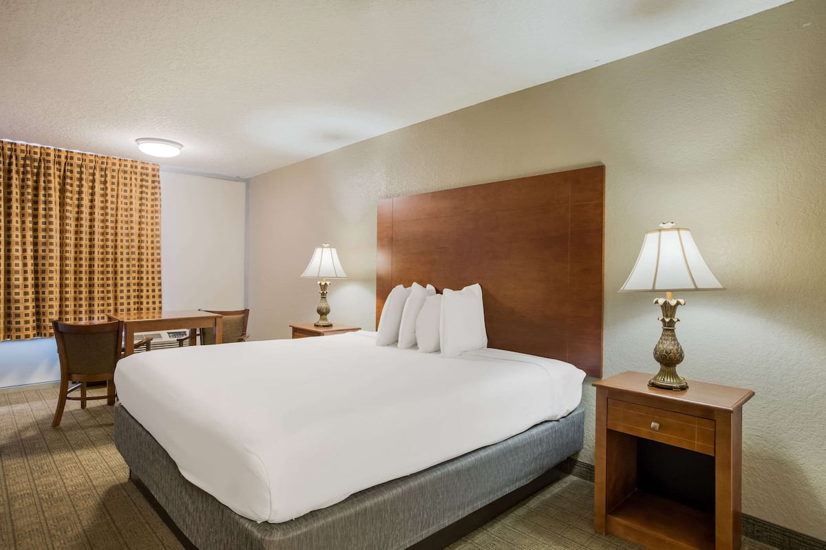 Quarto de hotel com uma cama grande, duas mesas de cabeceira com luminárias, uma escrivaninha com cadeira e uma janela com cortinas.