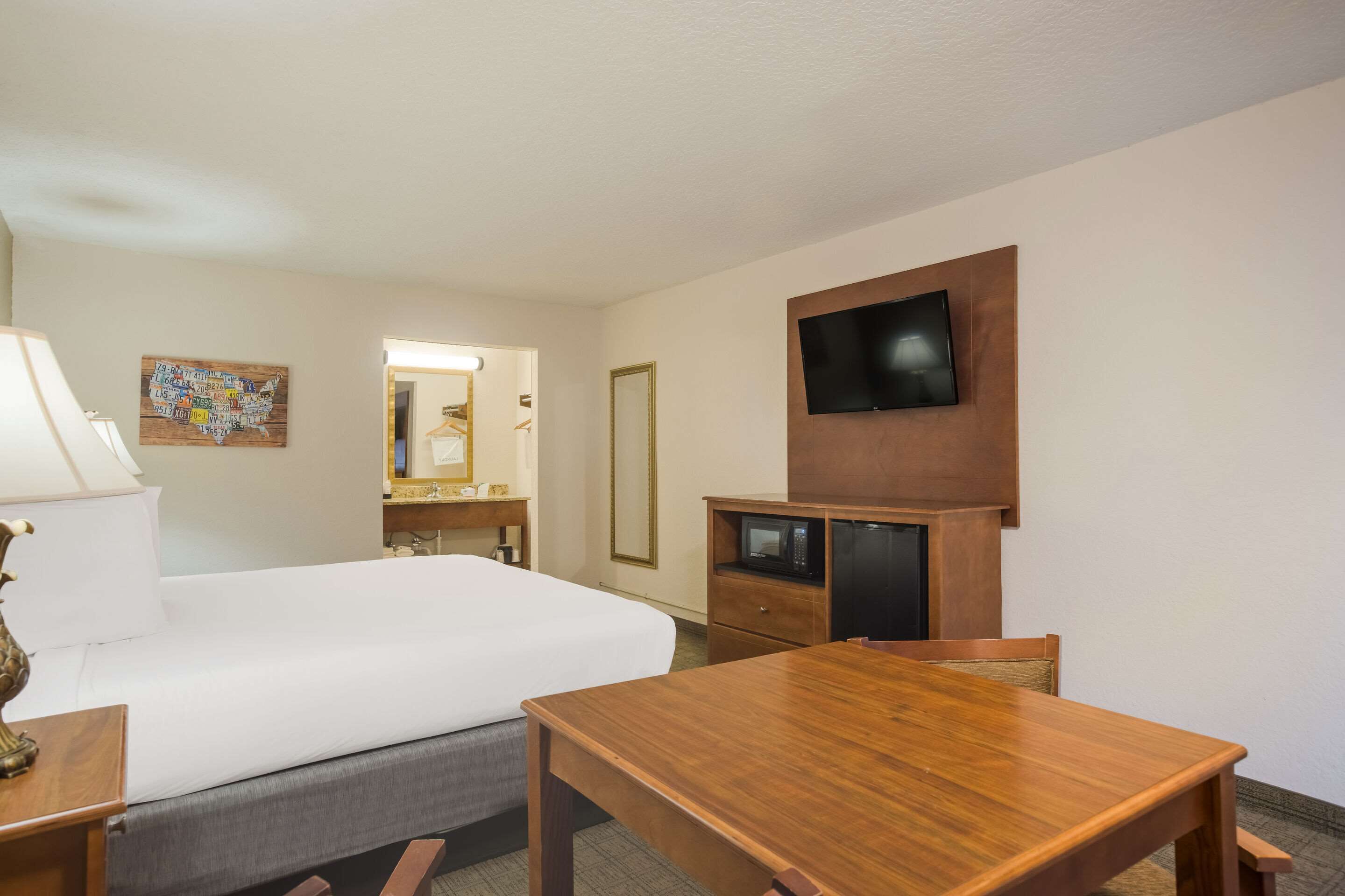 Quarto de hotel com cama, mesa de madeira, televisão fixada na parede, escrivaninha com luminária e espelho.