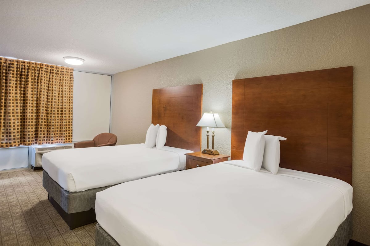Quarto de hotel com duas camas, uma escrivaninha, uma cadeira, um abajur e uma janela com cortinas.