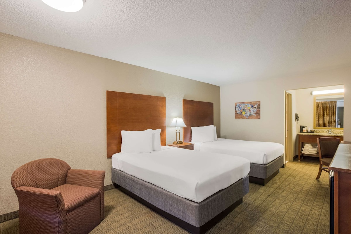 Quarto de hotel com duas camas, uma cadeira, uma escrivaninha e uma luminária.
