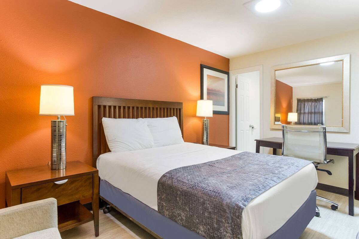 Quarto de hotel com cama, mesas de cabeceira com luminárias, escrivaninha com cadeira e um espelho grande.