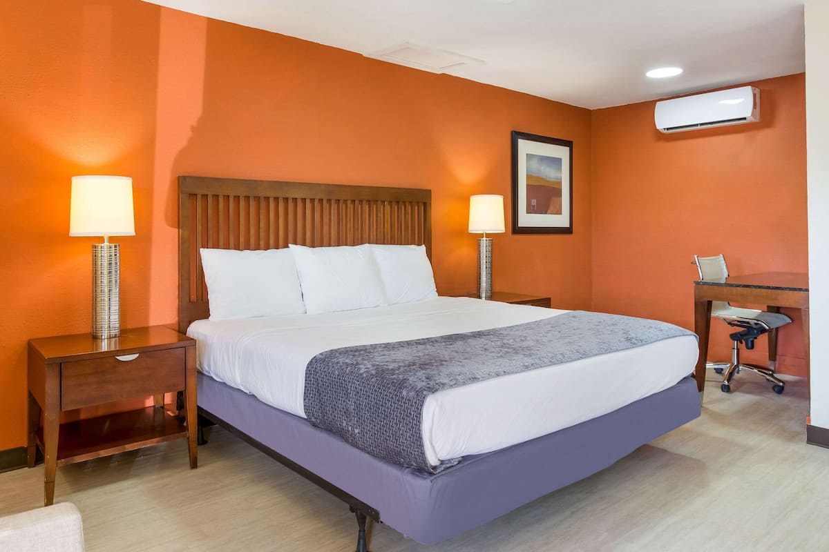 Quarto de hotel com cama, duas luminárias de apoio, escrivaninha com cadeira e um aparelho de ar condicionado na parede.
