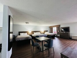 Quarto de hotel com cama, escrivaninha, duas cadeiras, televisão e janela com cortinas.