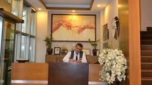 Uma mesa de recepção com um homem sentado atrás dela, um quadro grande acima e um vaso de flores à direita.