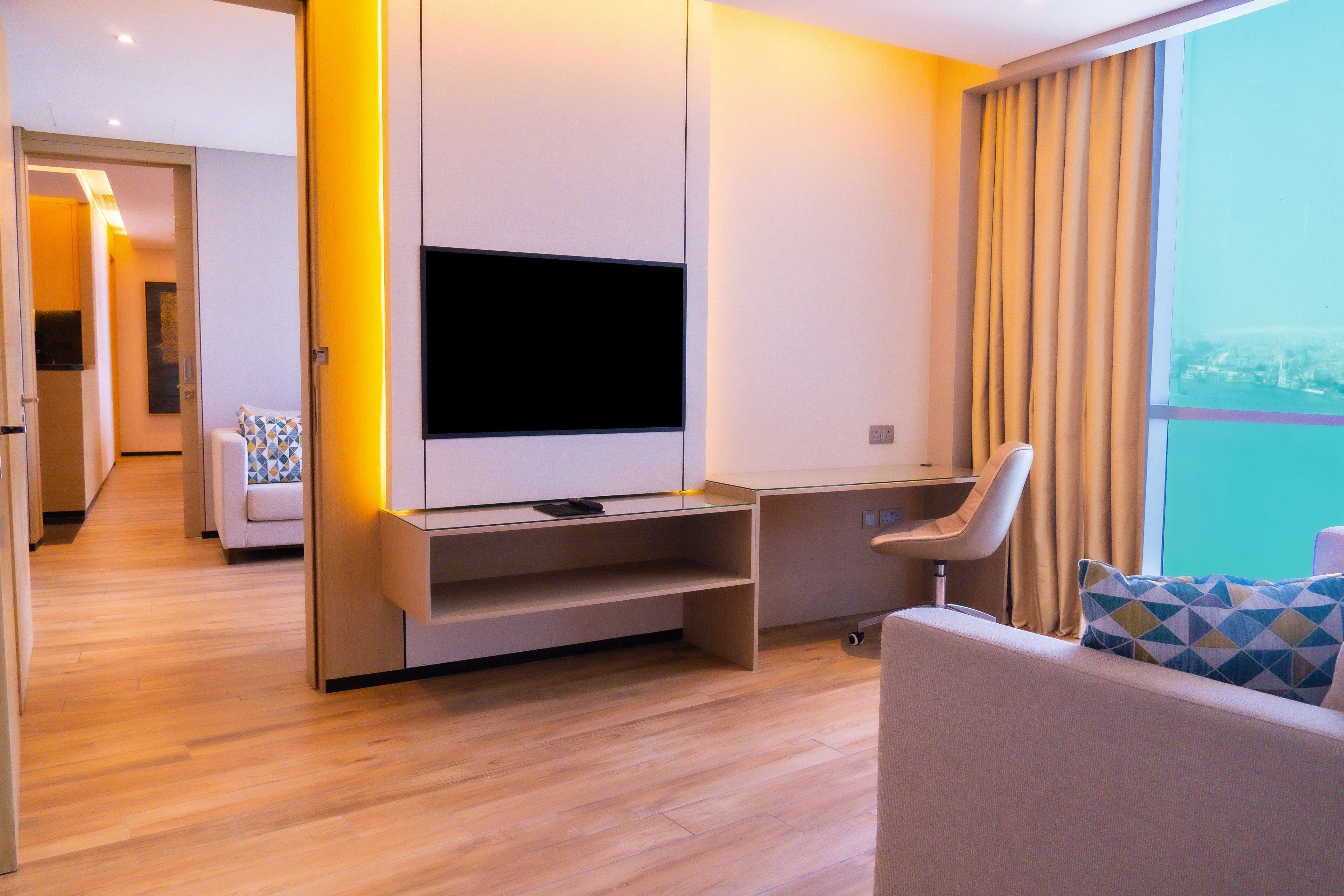 Quarto de hotel moderno com televisão de tela plana, sofá, escrivaninha e uma janela grande com cortinas.