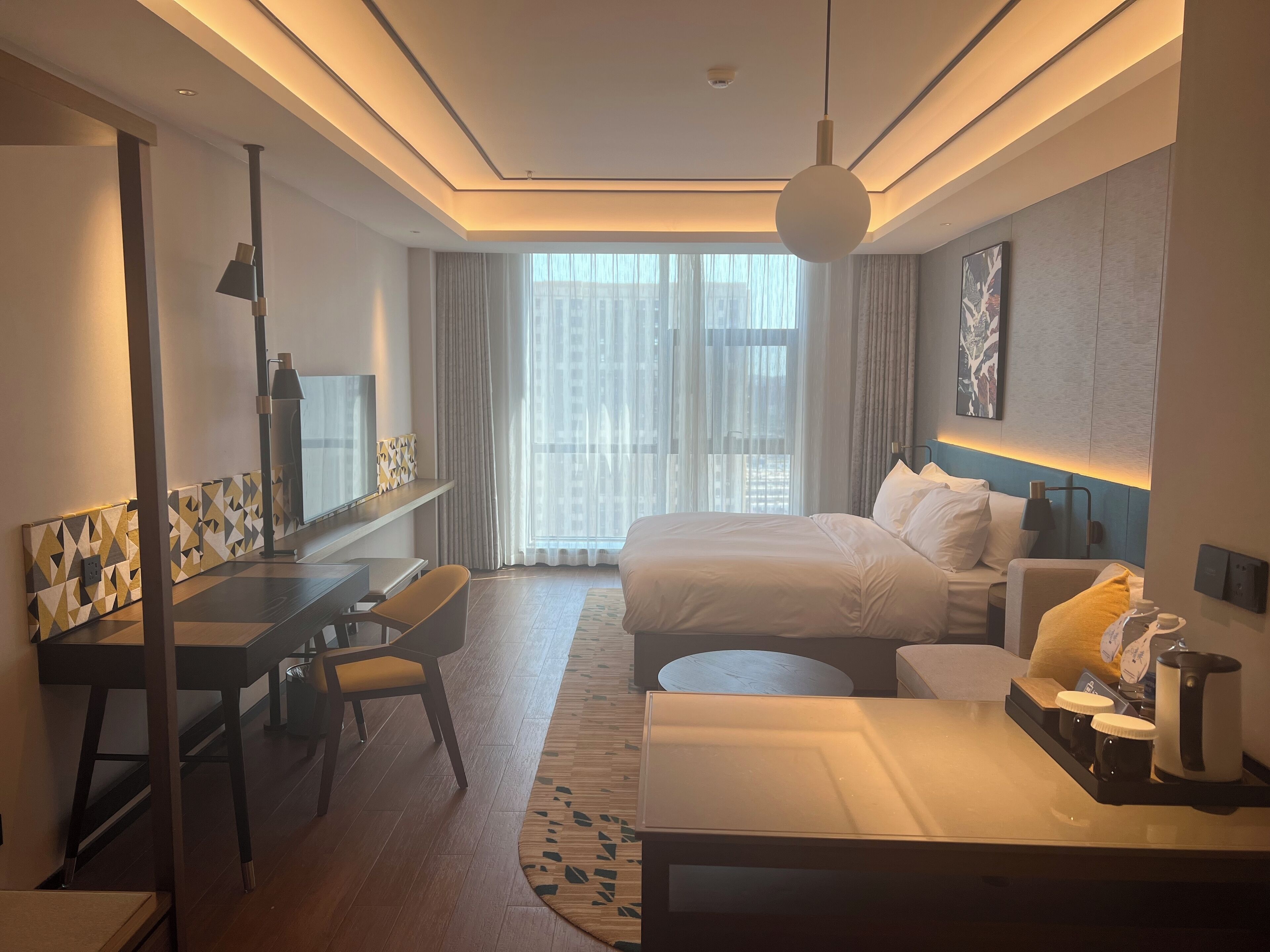 Quarto de hotel moderno com cama, escrivaninha, cadeira e uma grande janela com cortinas.