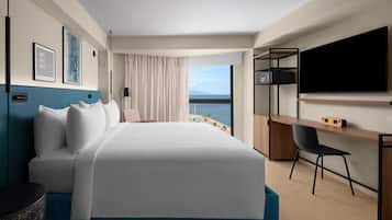 Quarto de hotel com cama, escrivaninha, cadeira, televisão e vista para o mar.