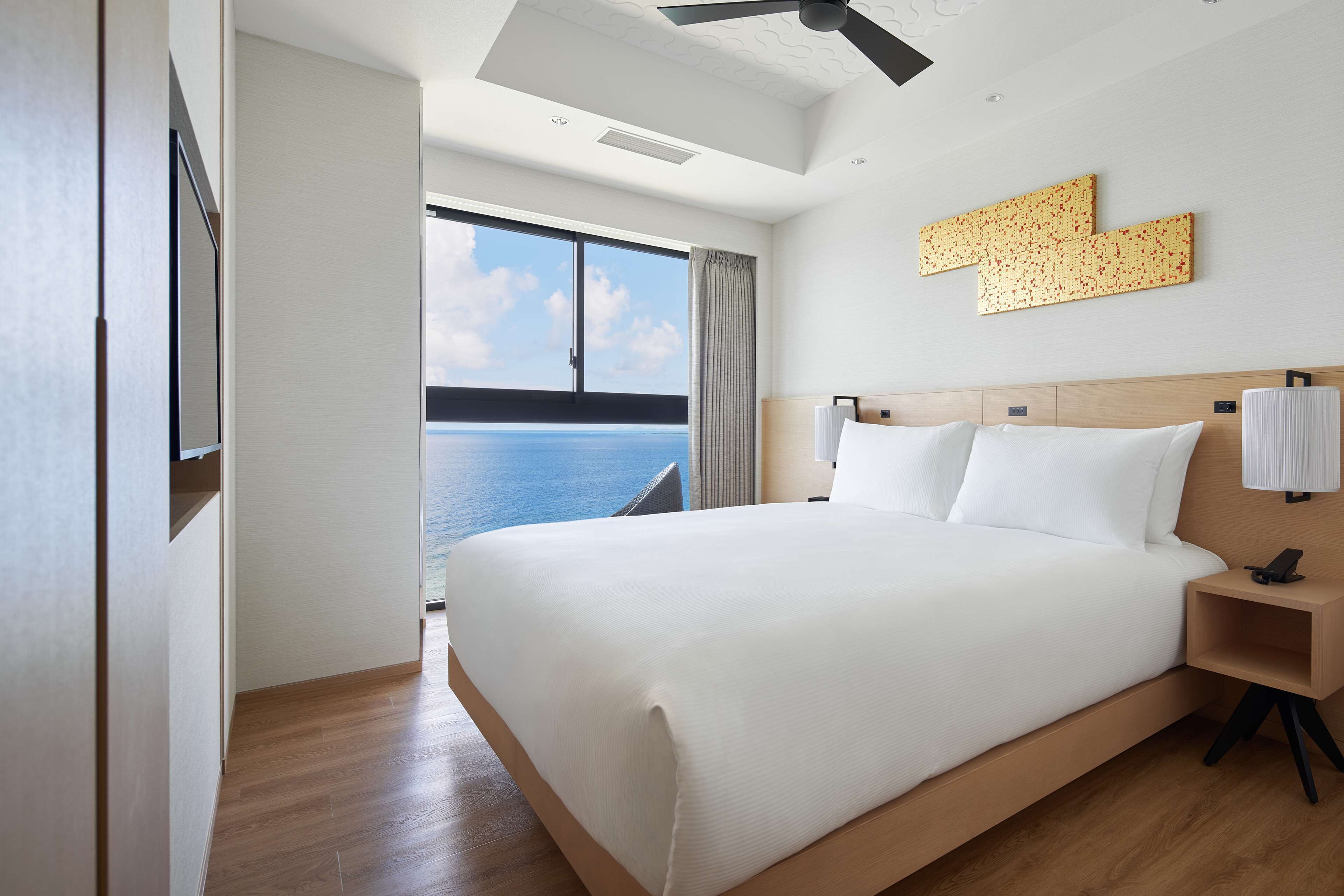 Quarto de hotel com cama grande, ventoinha no teto, vista para o mar e uma obra de arte na parede.