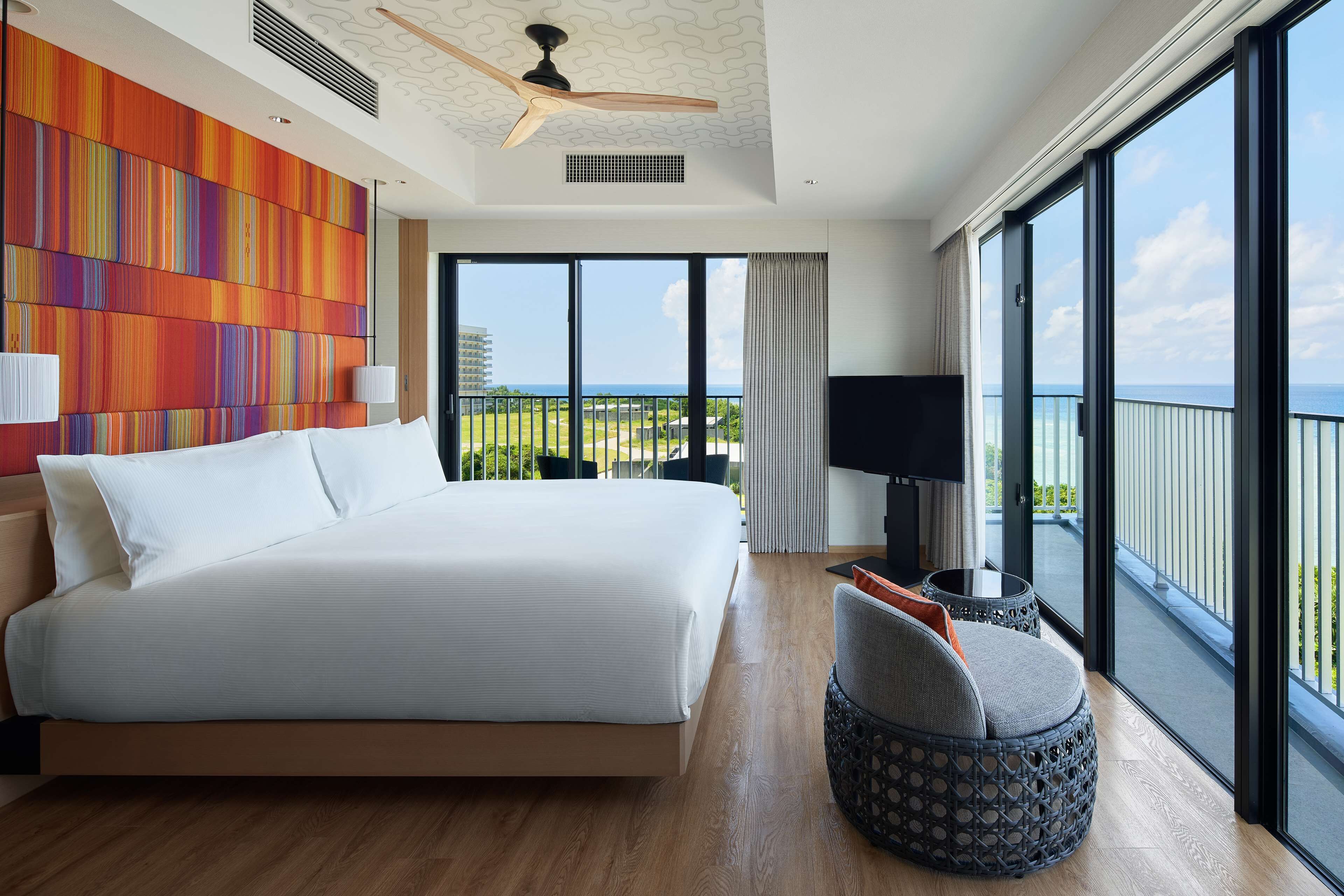 Quarto de hotel moderno com uma cama grande, televisão, varanda com vista para o oceano e ventilador de teto.