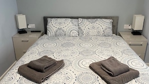 Cama bem arrumada com roupa de cama estampada, duas mesas de cabeceira com luminárias e um quadro pendurado acima da cama.